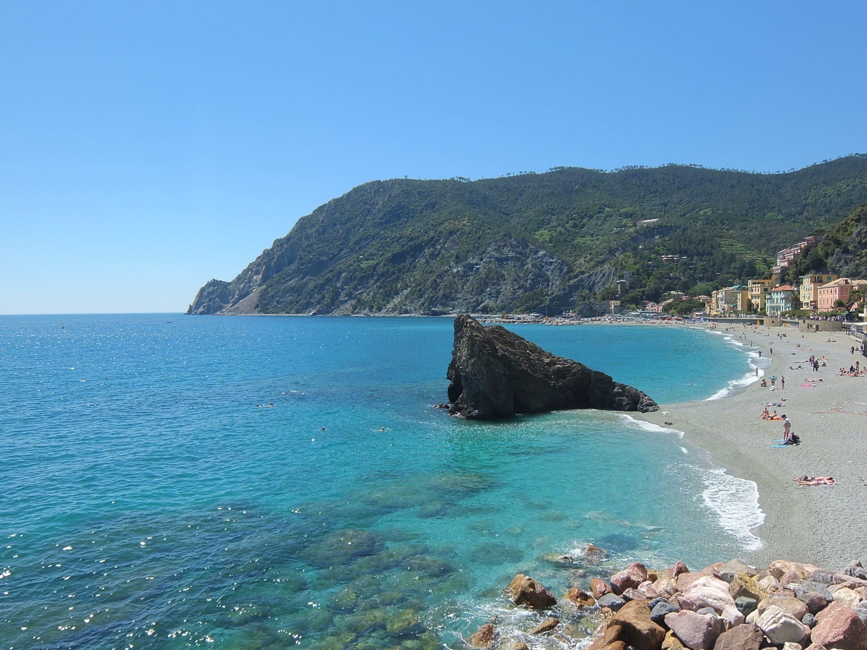 Uma praia costeira com águas turquesa cristalinas, uma formação rochosa imponente e um morro com edifícios.