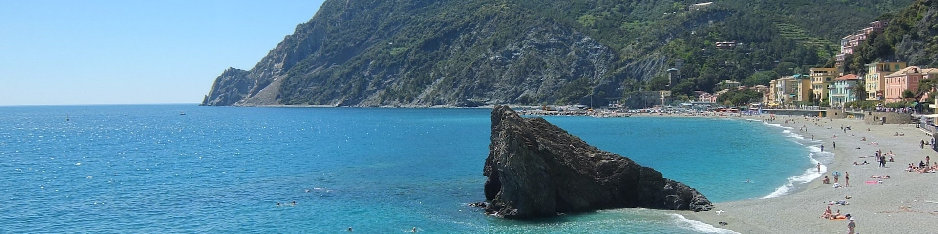 Uma praia costeira com águas turquesa cristalinas, uma formação rochosa imponente e um morro com edifícios.