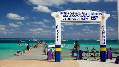 Um píer com pessoas e barcos, uma placa indicando 'Torre de Pesca Puerto Morelos' e uma placa da 'Corona'.