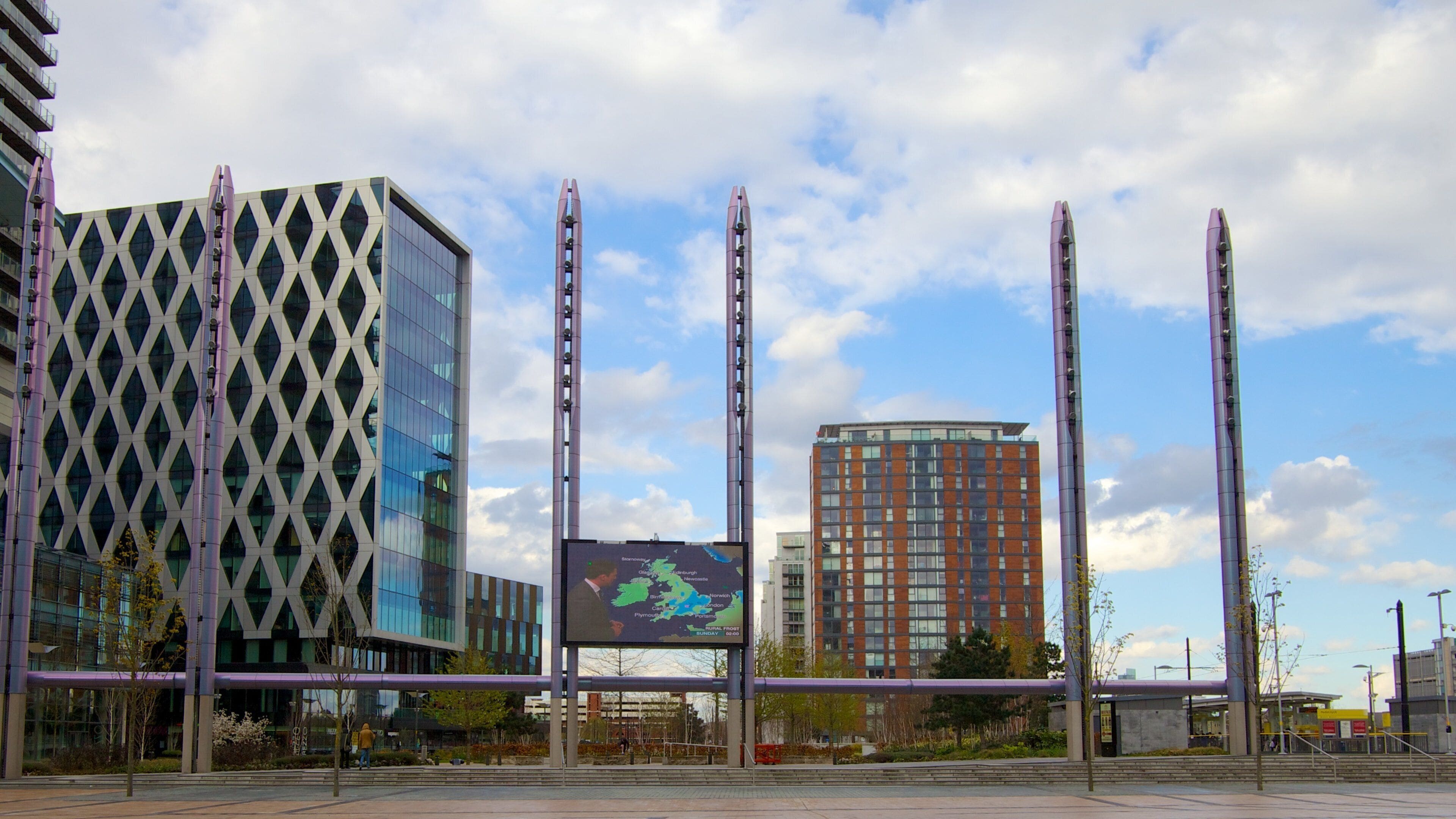 Uma praça urbana moderna com edifícios contemporâneos, um grande painel digital e céu claro.