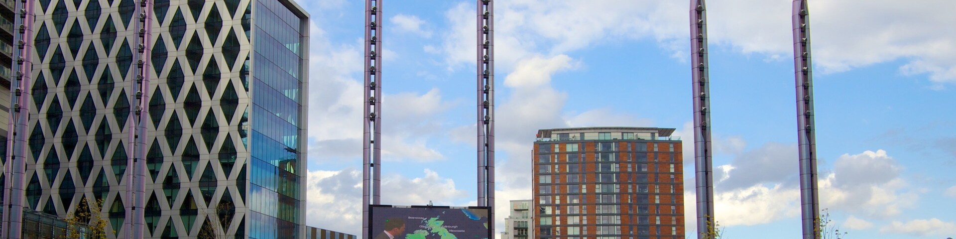 Uma praça urbana moderna com edifícios contemporâneos, um grande painel digital e céu claro.