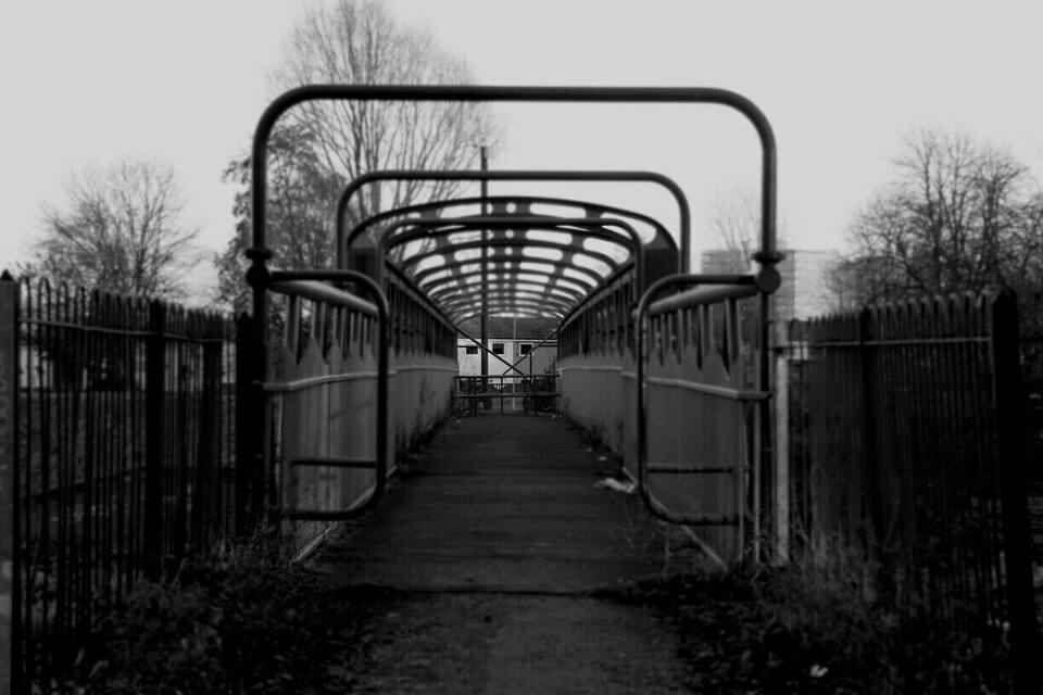 Uma ponte de pedestres de metal com um telhado curvo, rodeada por uma cerca de metal e árvores.