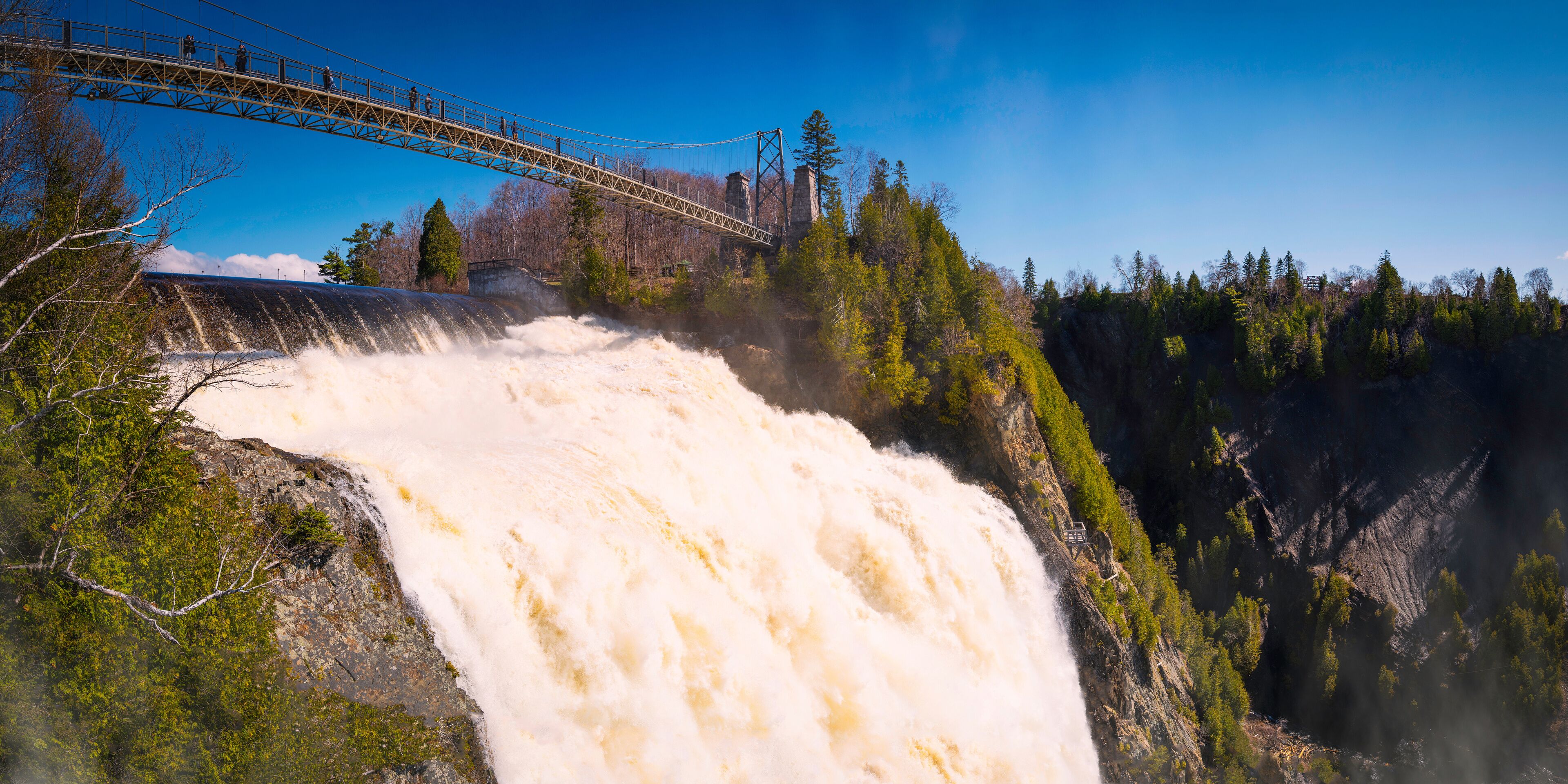 Uma grande cascata desce por um penhasco rochoso, uma ponte suspensa e uma floresta densa ao fundo.