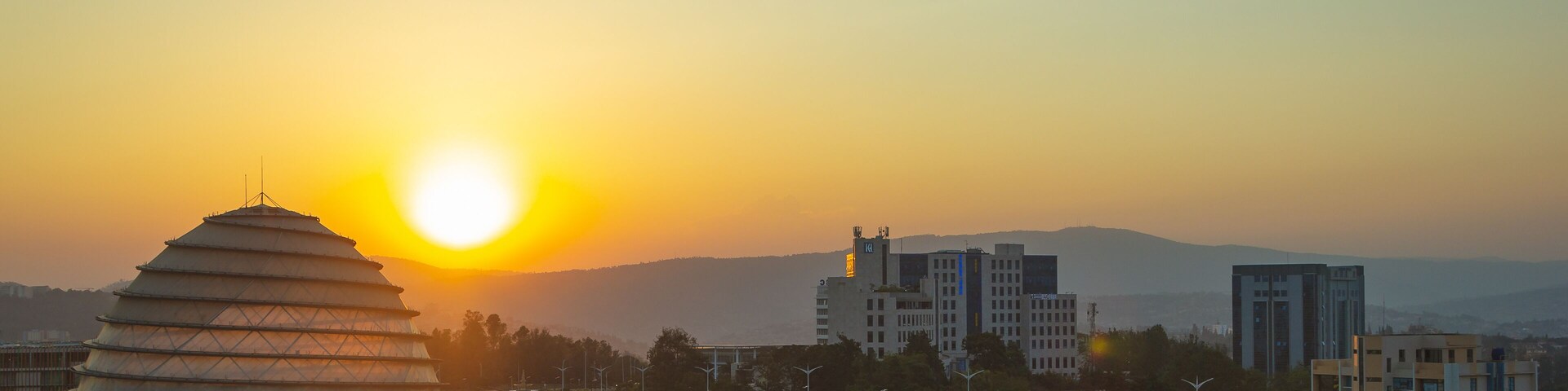 Pôr do sol sobre uma paisagem urbana, com uma estrutura semelhante a um pagode em primeiro plano e edifícios modernos ao fundo.