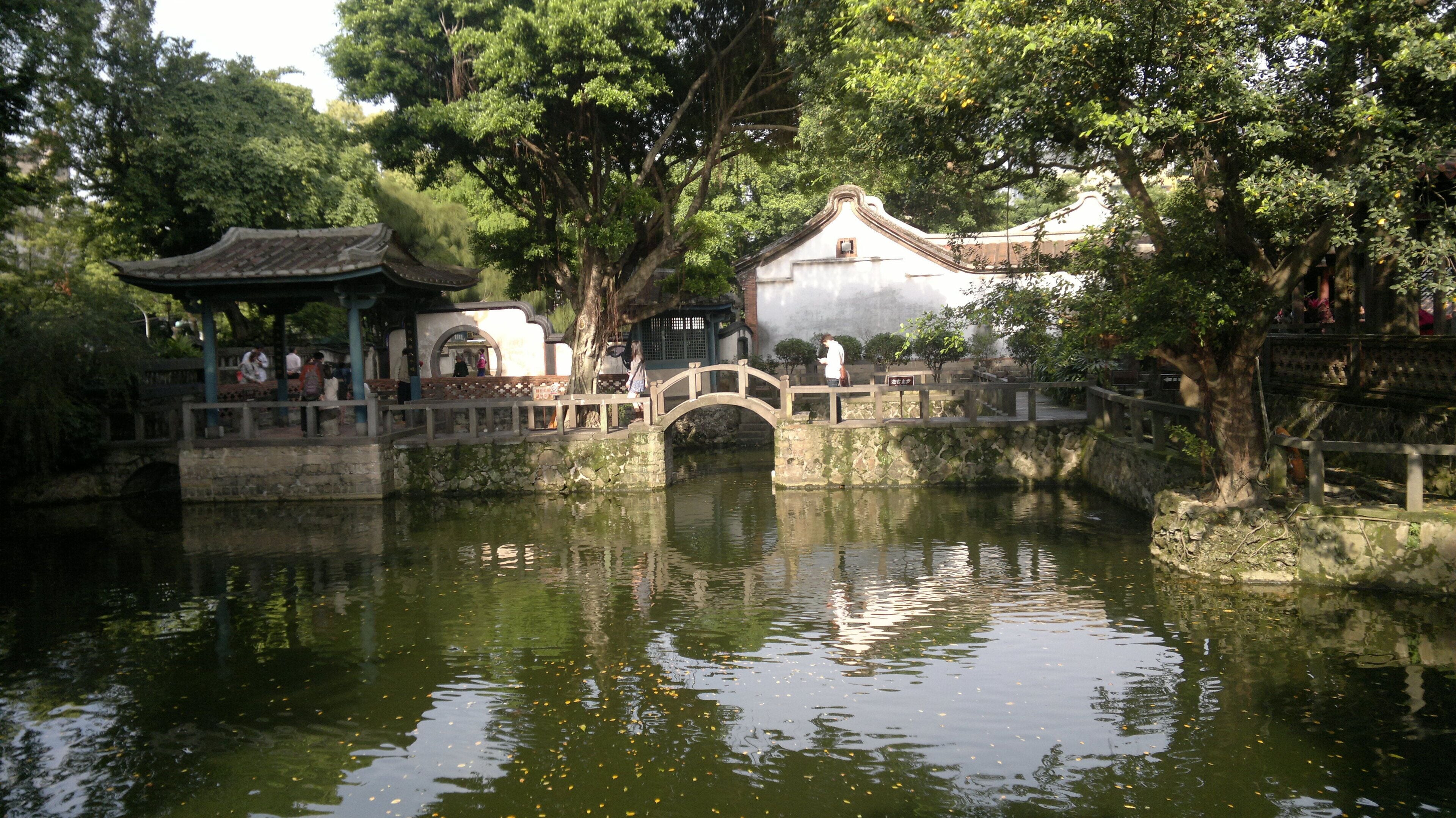 Um jardim tradicional chinês com uma ponte de pedra sobre um lago, rodeado por árvores exuberantes e um edifício branco com arquitetura ornamentada.