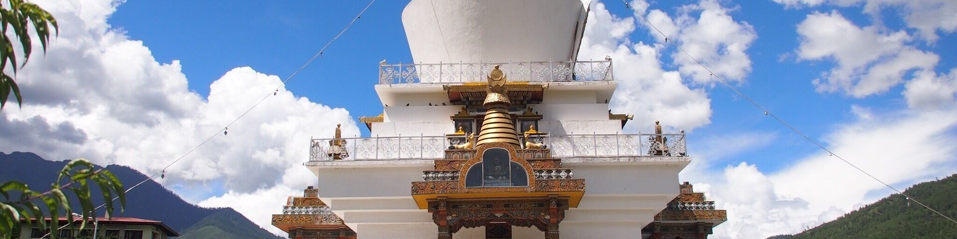 Um stupa branco com detalhes dourados, rodeado por vegetação e montanhas.