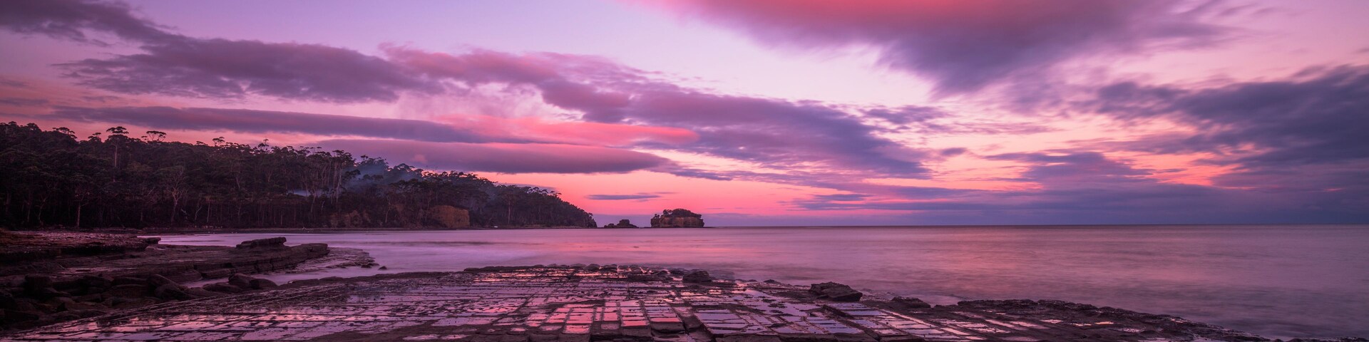 Paisagem costeira com céu ao pôr do sol, linha de costa rochosa e mar calmo.