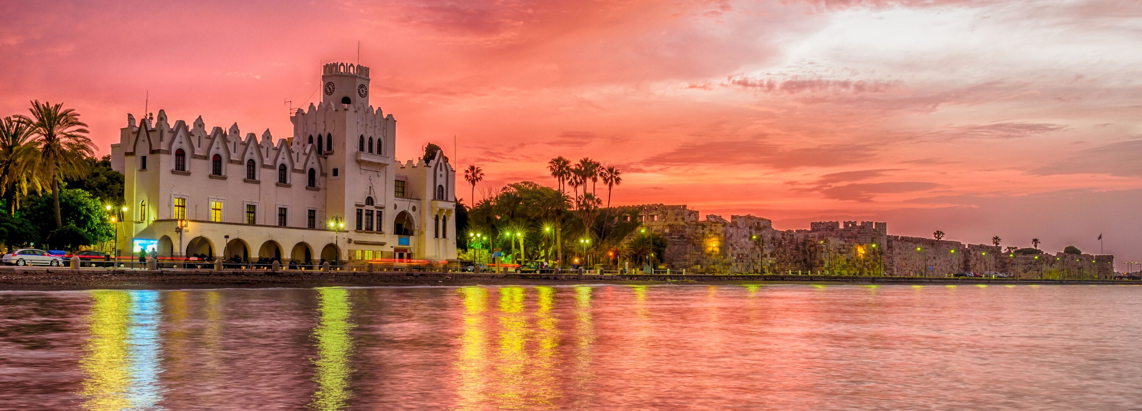 Um edifício histórico com uma torre do relógio, palmeiras e uma orla ao pôr do sol.