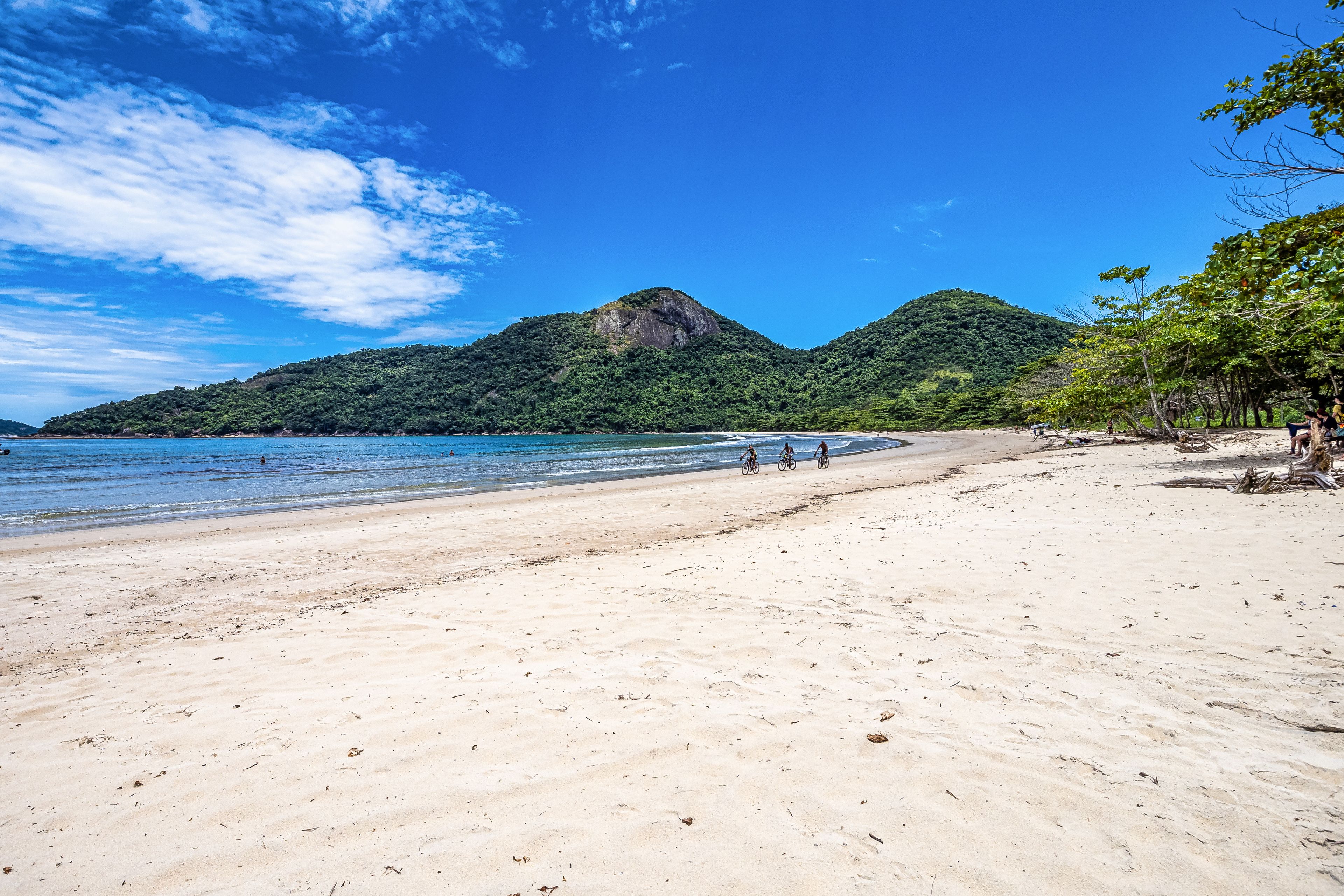 Uma praia com águas azul-claras, areal arenoso e colinas verdejantes ao fundo.