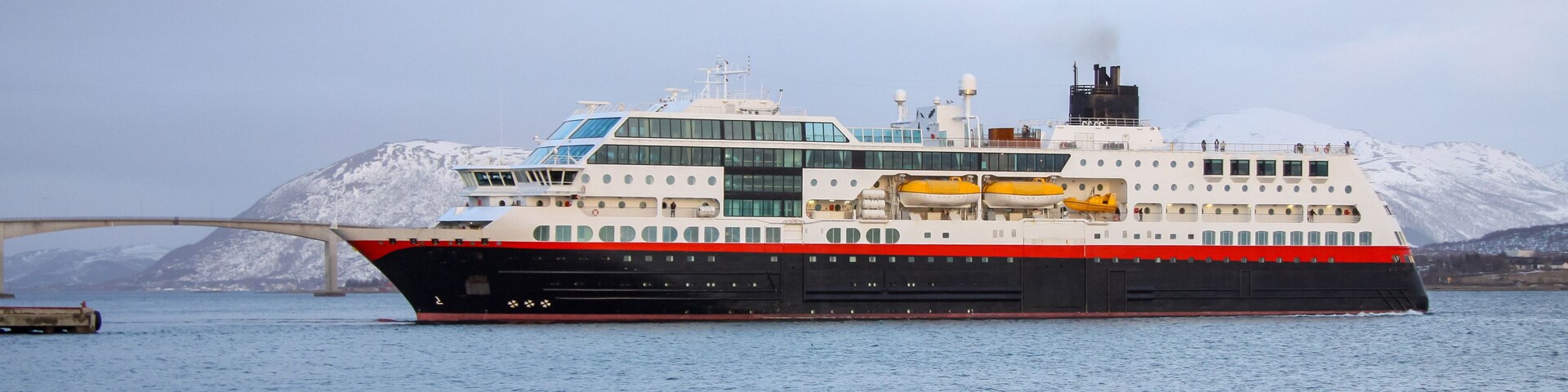 Um grande navio de cruzeiro com casco vermelho e preto, superestrutura branca e botes salva-vidas amarelos, atracado perto de uma ponte e montanhas cobertas de neve.