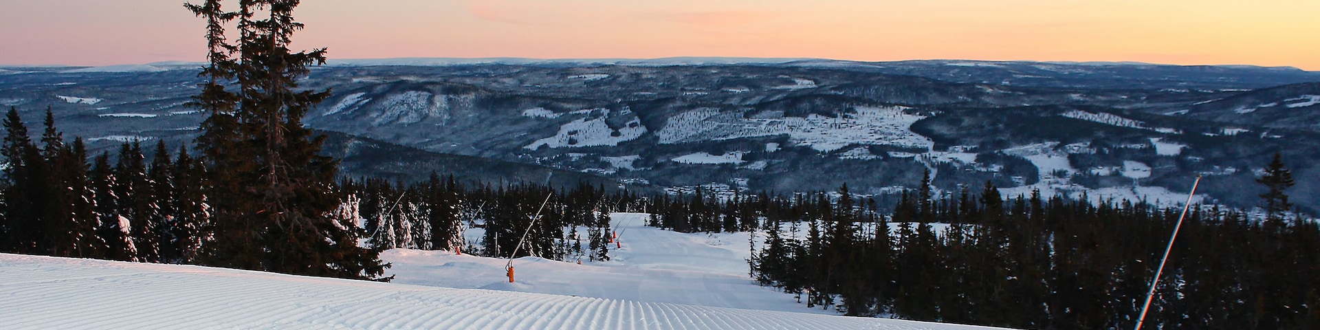 Paisagem montanhosa coberta de neve com uma pista de esqui, árvores e um pôr do sol ao fundo.