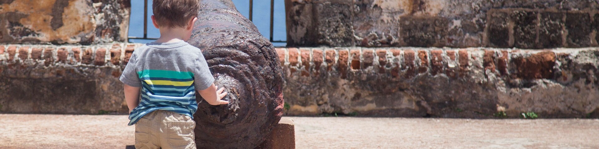 Criança em frente a uma estátua grande e desgastada, com uma janela gradeada azul ao fundo.