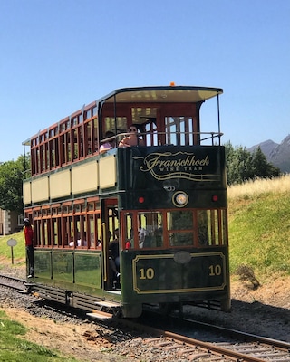 Um elétrico de dois andares antigo com a inscrição 'Franschhoek Wine Tram', com passageiros, percorrendo trilhos por uma paisagem rural com montanhas ao fundo.
