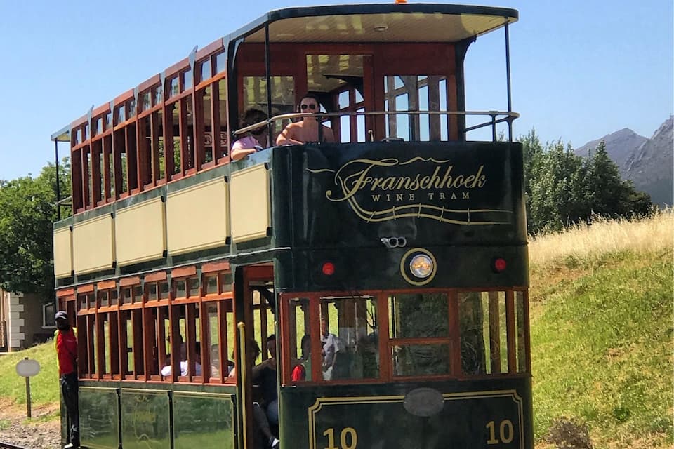 Um elétrico de dois andares antigo com a inscrição 'Franschhoek Wine Tram', com passageiros, percorrendo trilhos por uma paisagem rural com montanhas ao fundo.