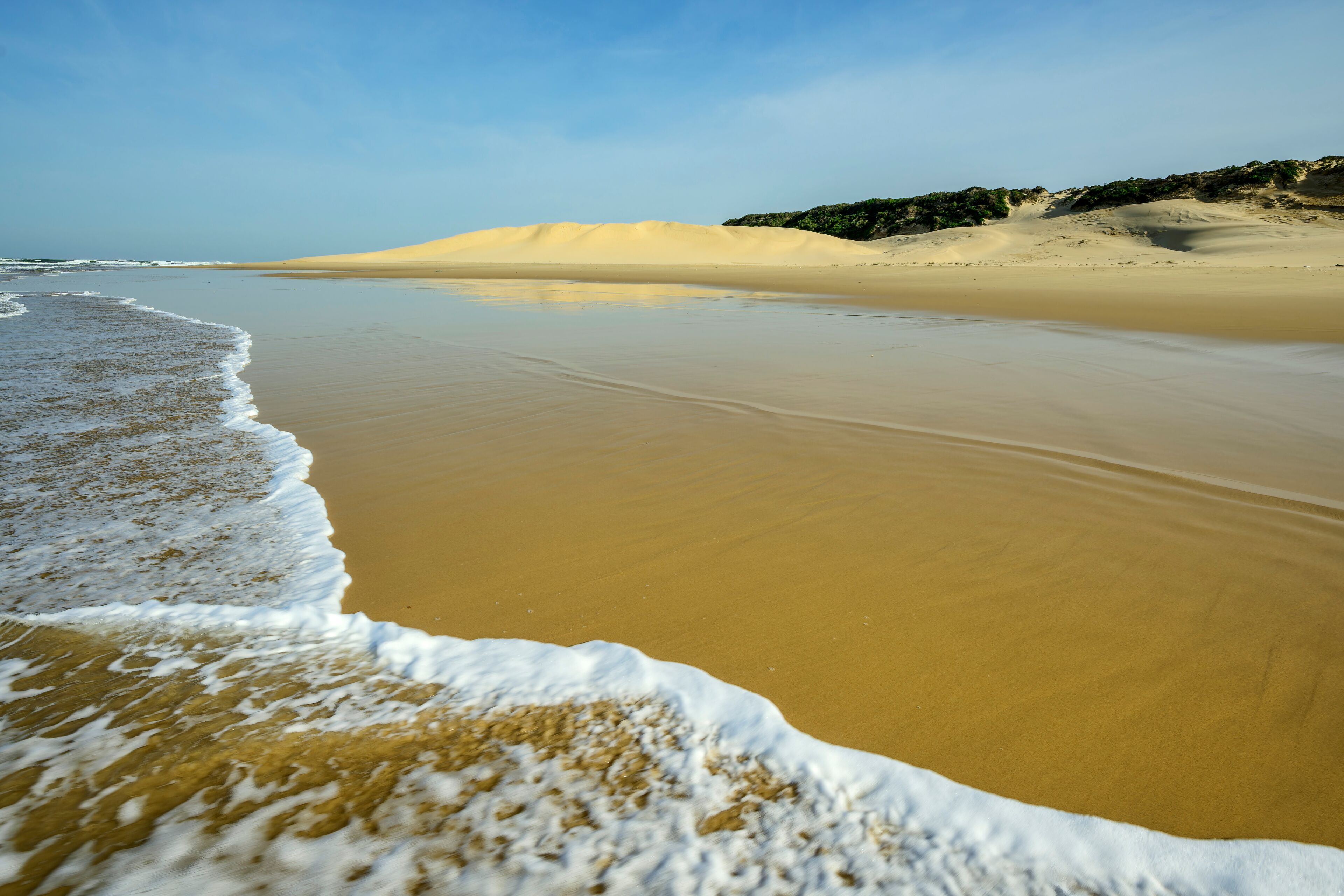 Uma praia arenosa com ondas suaves, céu claro e dunas ao longe.
