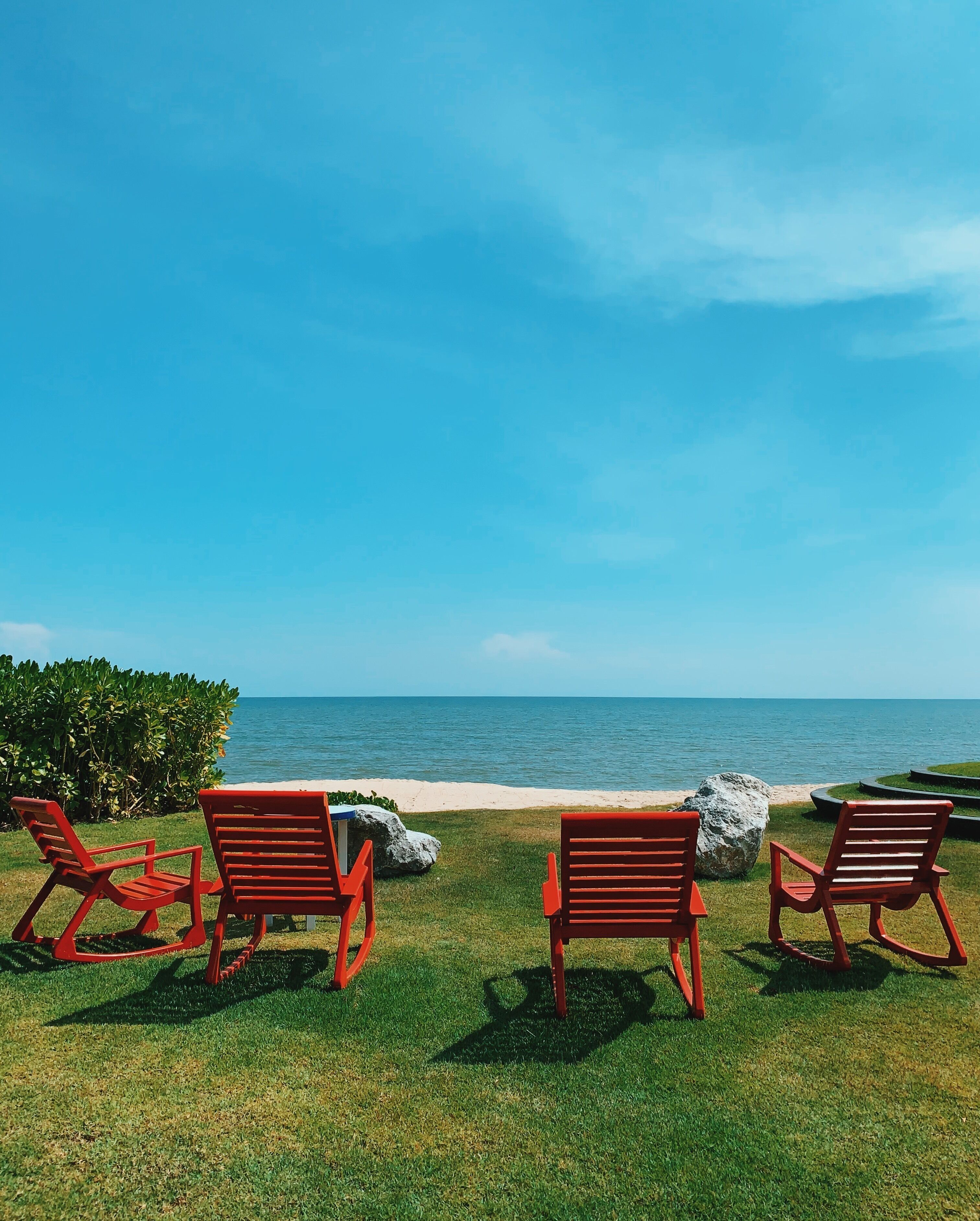Três poltronas vermelhas voltadas para uma praia, com céu azul e límpido.