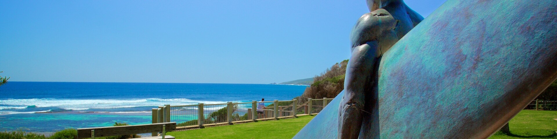 Estátua de bronze de um homem segurando uma prancha de surf, com vista para uma praia com ondas e céu azul claro.