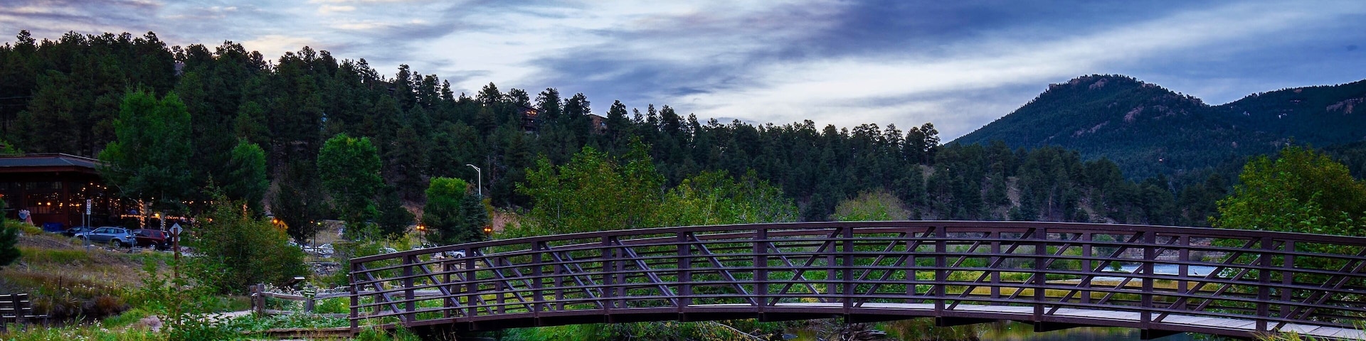 Uma ponte de madeira sobre um rio calmo, cercada por vegetação exuberante e uma encosta arborizada.