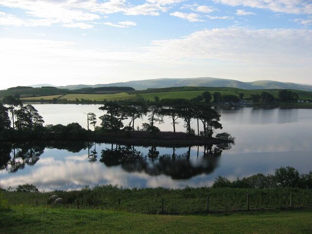 Um lago tranquilo com uma ilha arborizada, colinas suaves ao fundo e céu limpo.