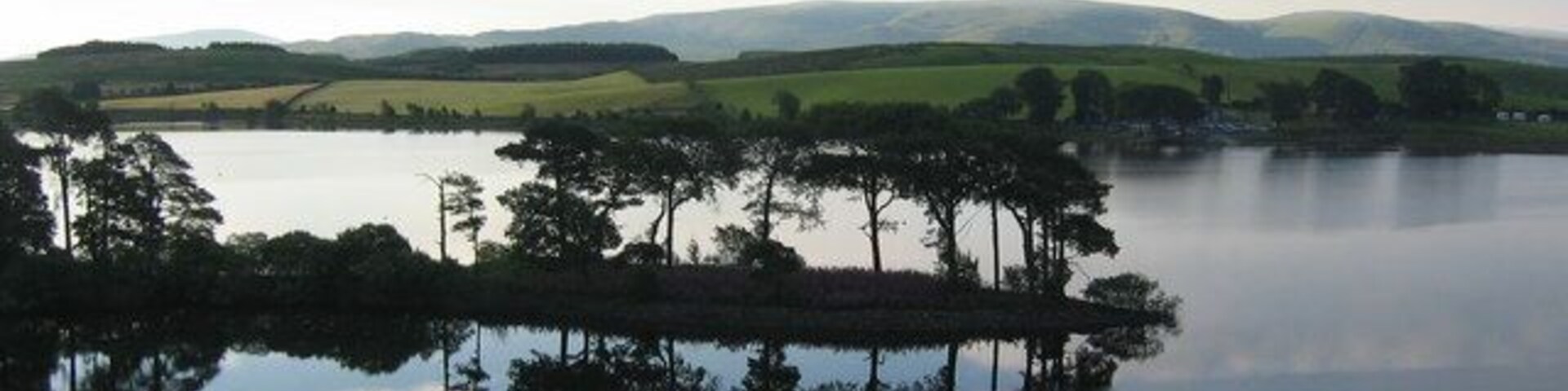 Um lago tranquilo com uma ilha arborizada, colinas suaves ao fundo e céu limpo.