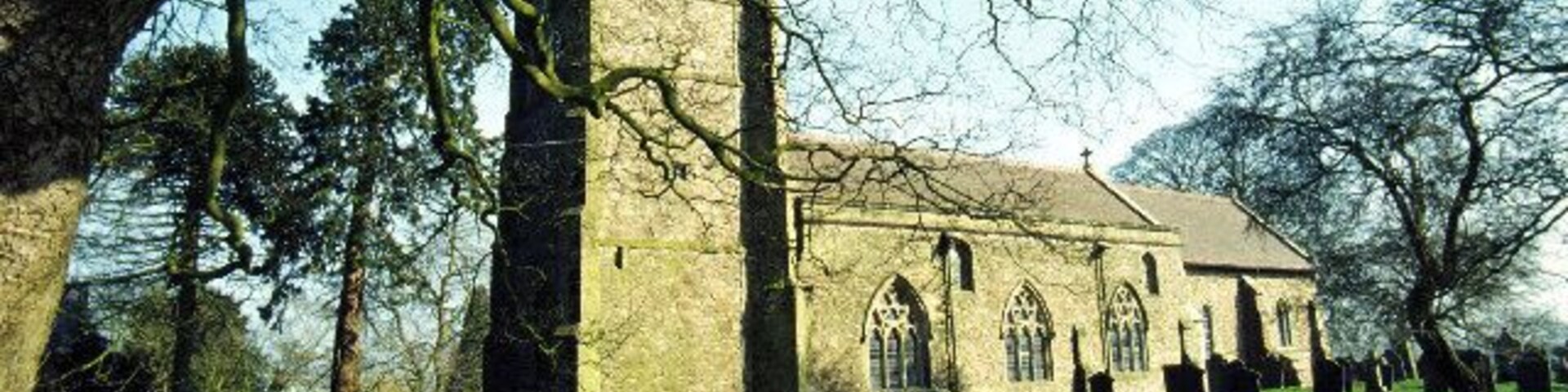 Igreja com uma torre alta e janelas em arco, cercada por um campo gramado e árvores.