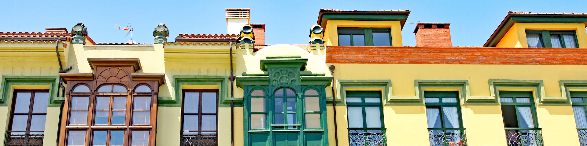 Uma fileira de edifícios coloridos com estilos arquitetônicos distintos, incluindo fachadas verdes e marrons e varandas ornamentadas.