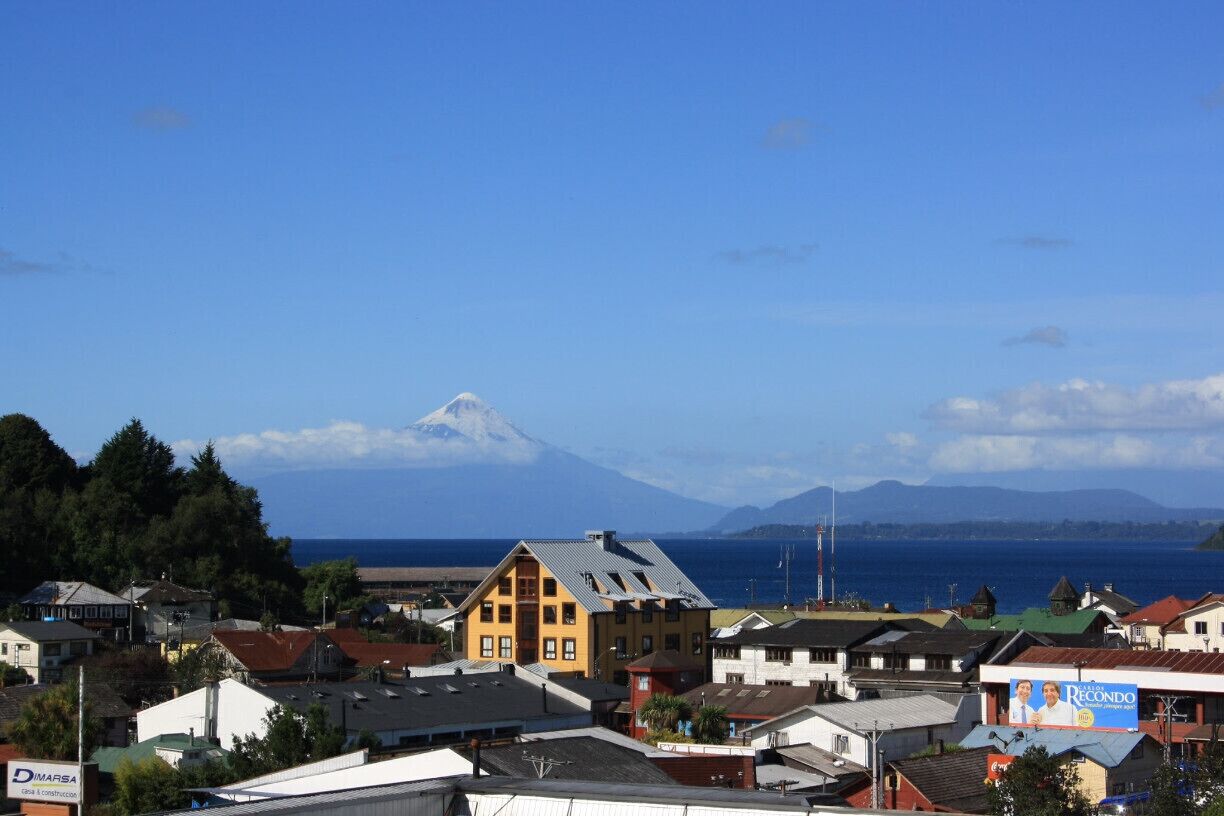 Uma cidade costeira com casas coloridas, um edifício amarelo em destaque e uma montanha ao fundo.