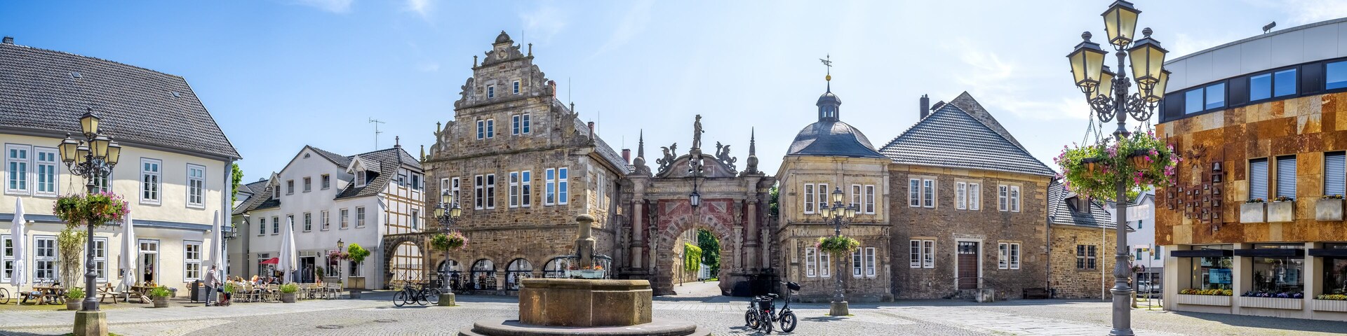 Uma praça de paralelepípedos com edifícios históricos, uma fonte e áreas de estar ao ar livre.