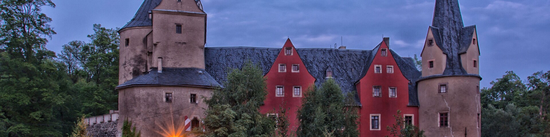 Um castelo histórico com várias torres e uma torre central imponente, rodeado por vegetação exuberante e um caminho de pedras.