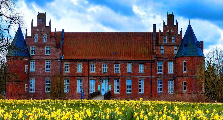Um grande edifício de tijolos vermelhos com várias torres e um campo de flores amarelas na frente.
