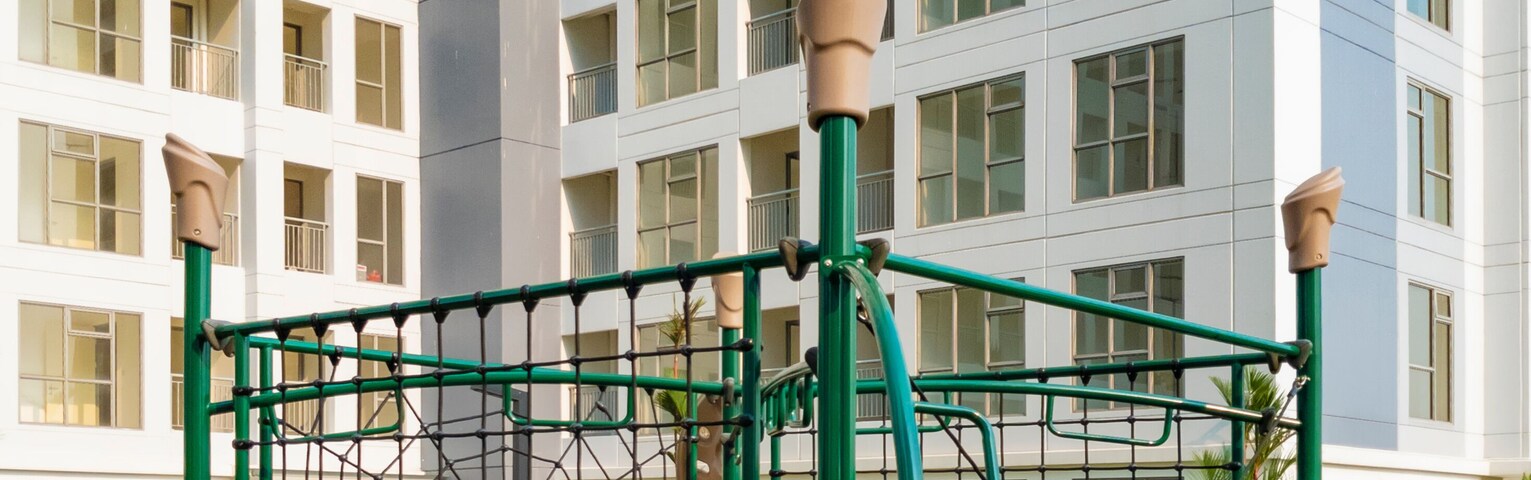 Um parquinho com escorrega e rede de escalada, cercado por um prédio com várias janelas.