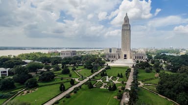 Um edifício grande e histórico com uma torre alta, cercado por jardins bem cuidados e um rio ao fundo.