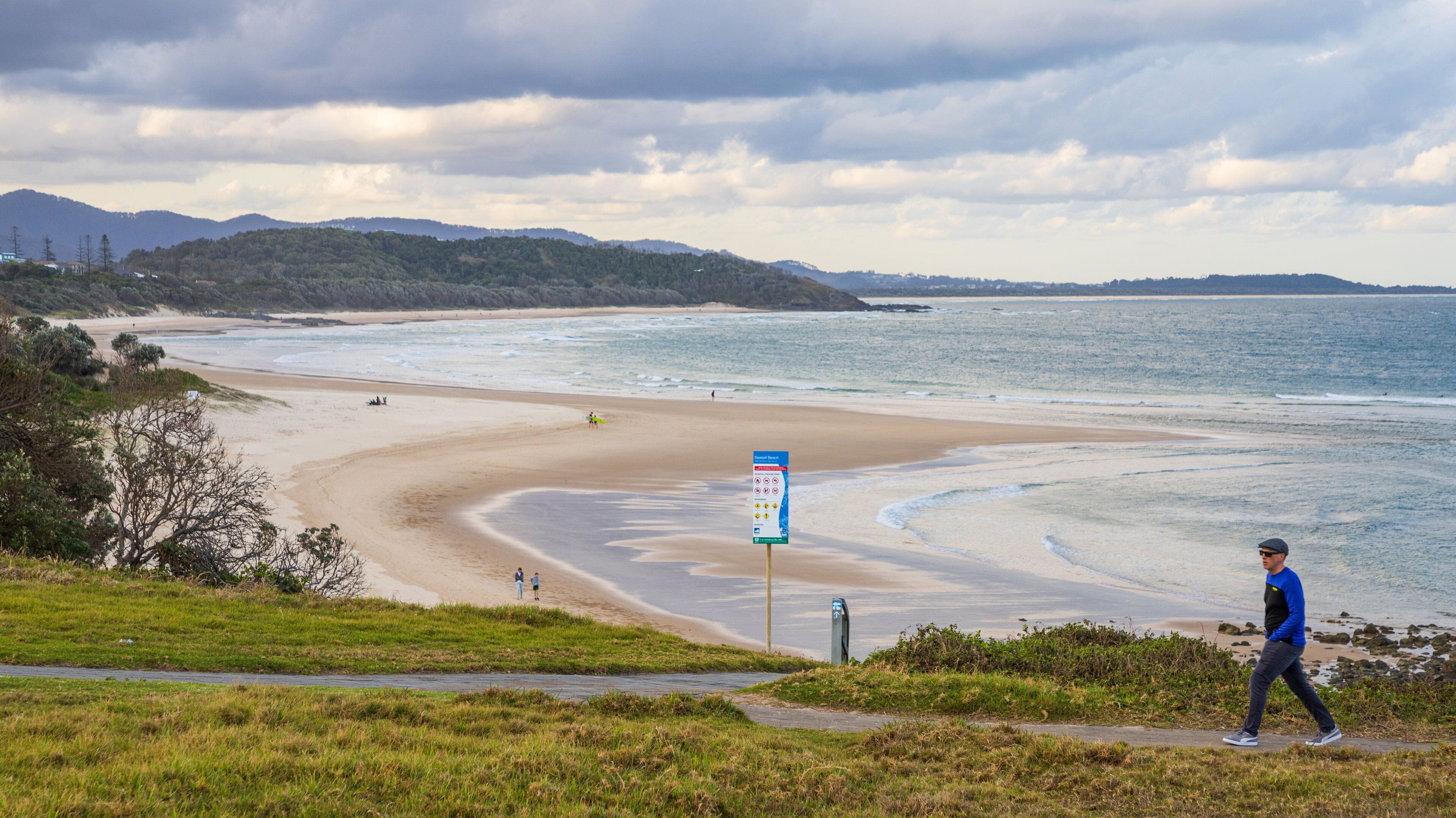 Uma praia com uma placa, uma pessoa caminhando e uma paisagem costeira com colinas ao fundo.