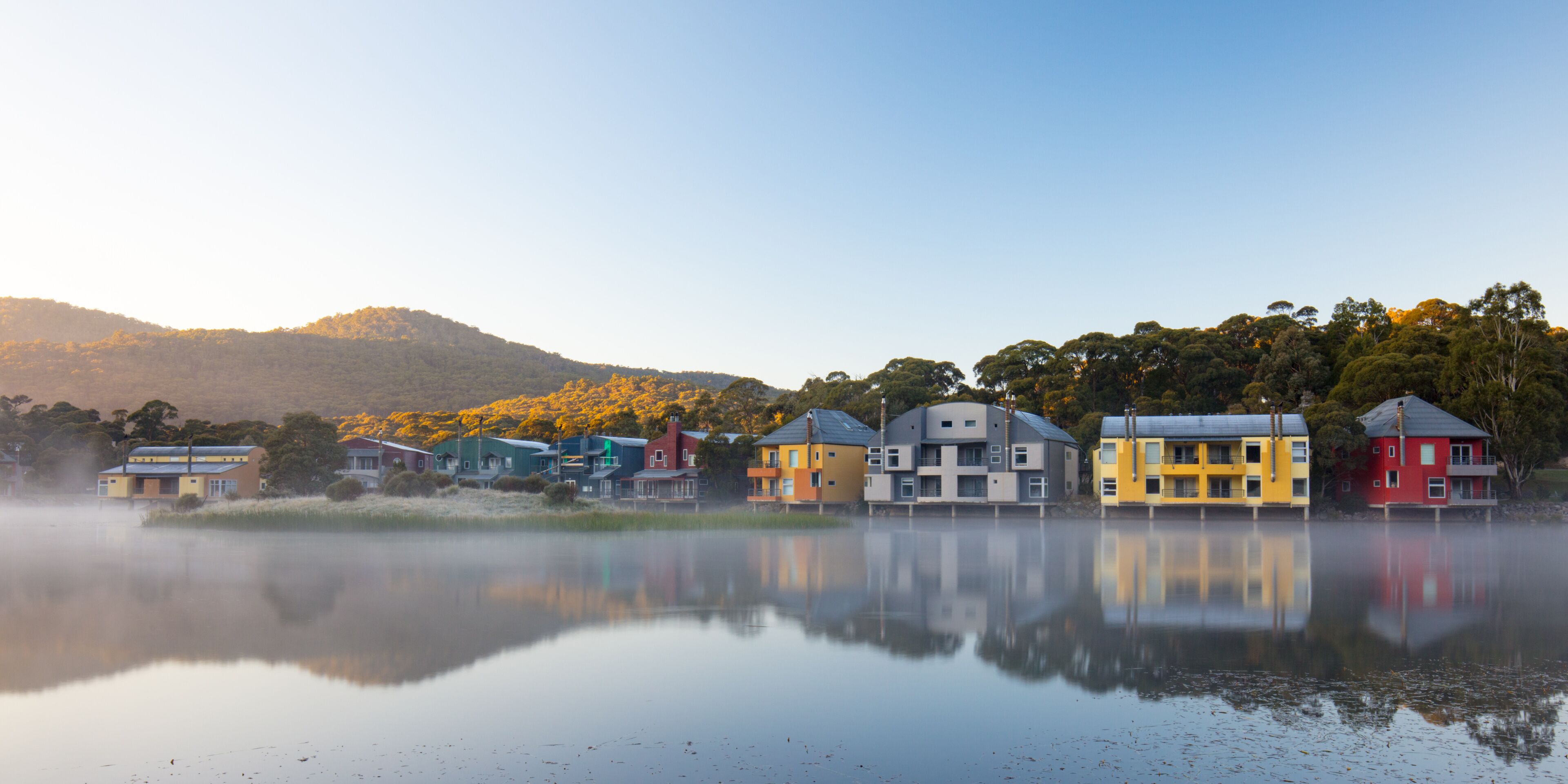 Um lago calmo com casas coloridas refletidas na água, cercado por árvores e colinas.