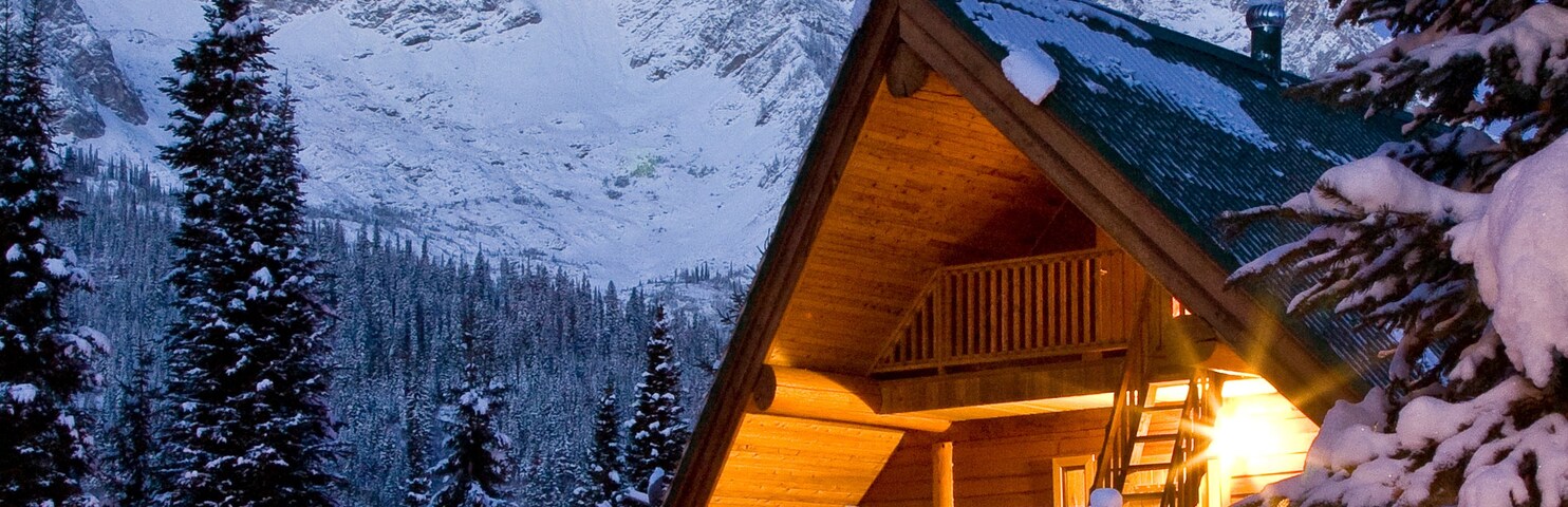 Uma cabana de madeira com varanda, rodeada por árvores cobertas de neve e montanhas ao fundo.
