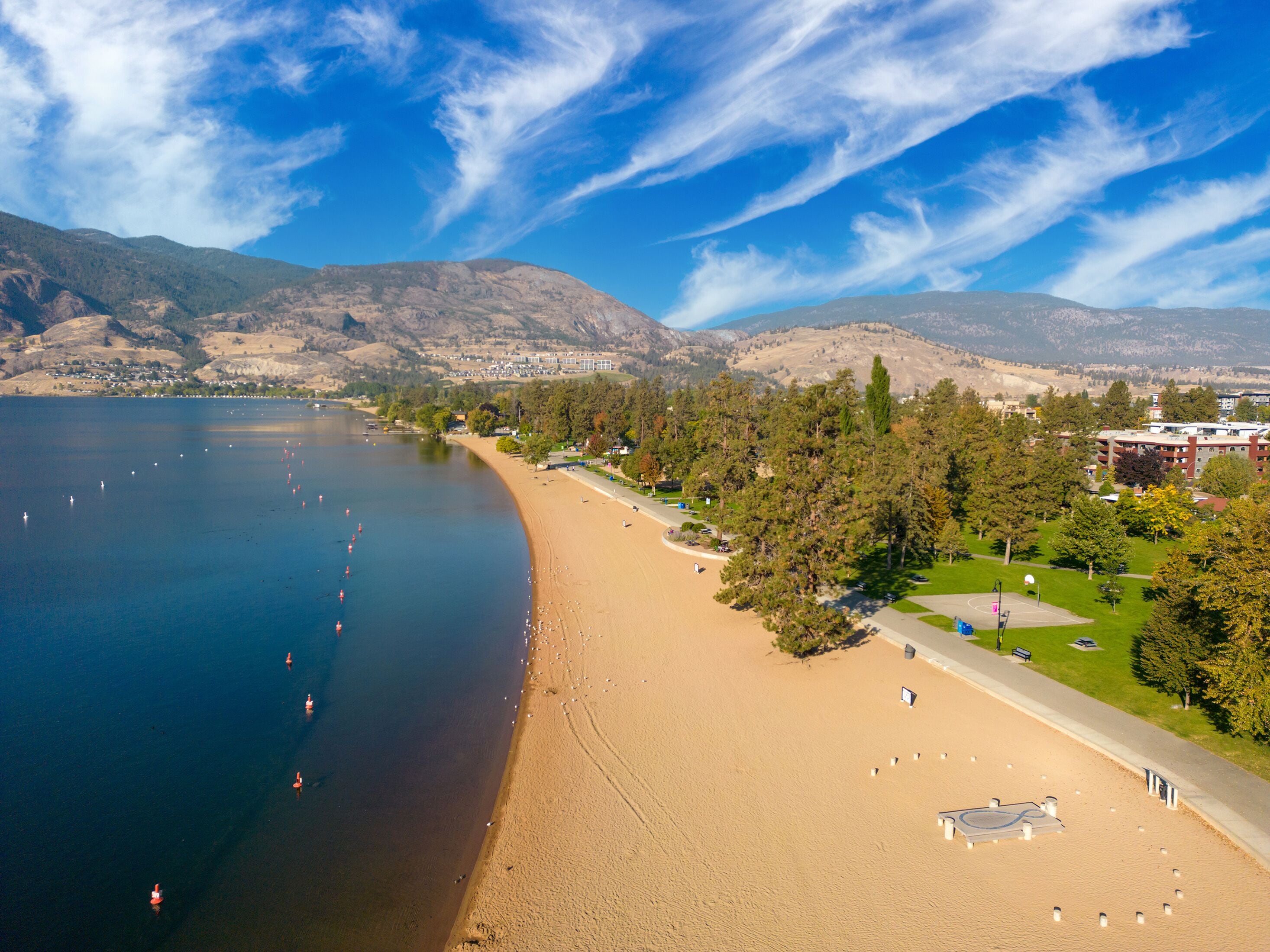 Paisagem costeira com praia arenosa, lago calmo e colinas e edifícios ao fundo.