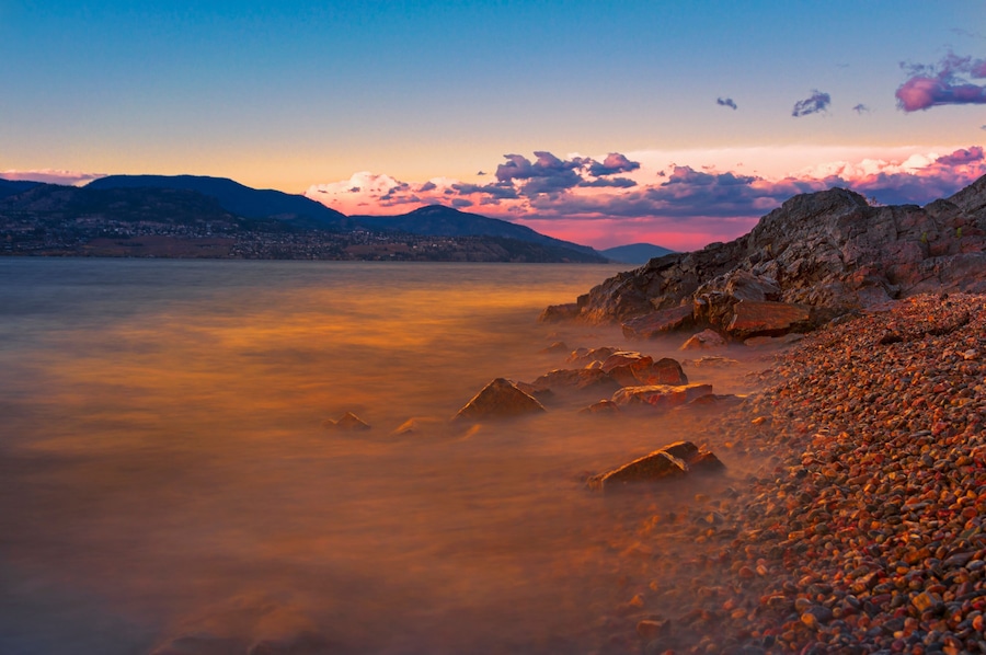 Costa rochosa com um lago calmo e montanhas ao longe durante o pôr do sol.