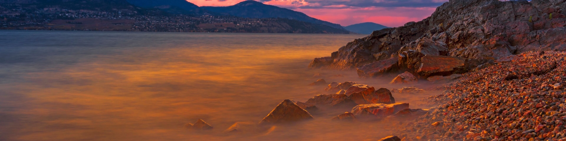 Costa rochosa com um lago calmo e montanhas ao longe durante o pôr do sol.