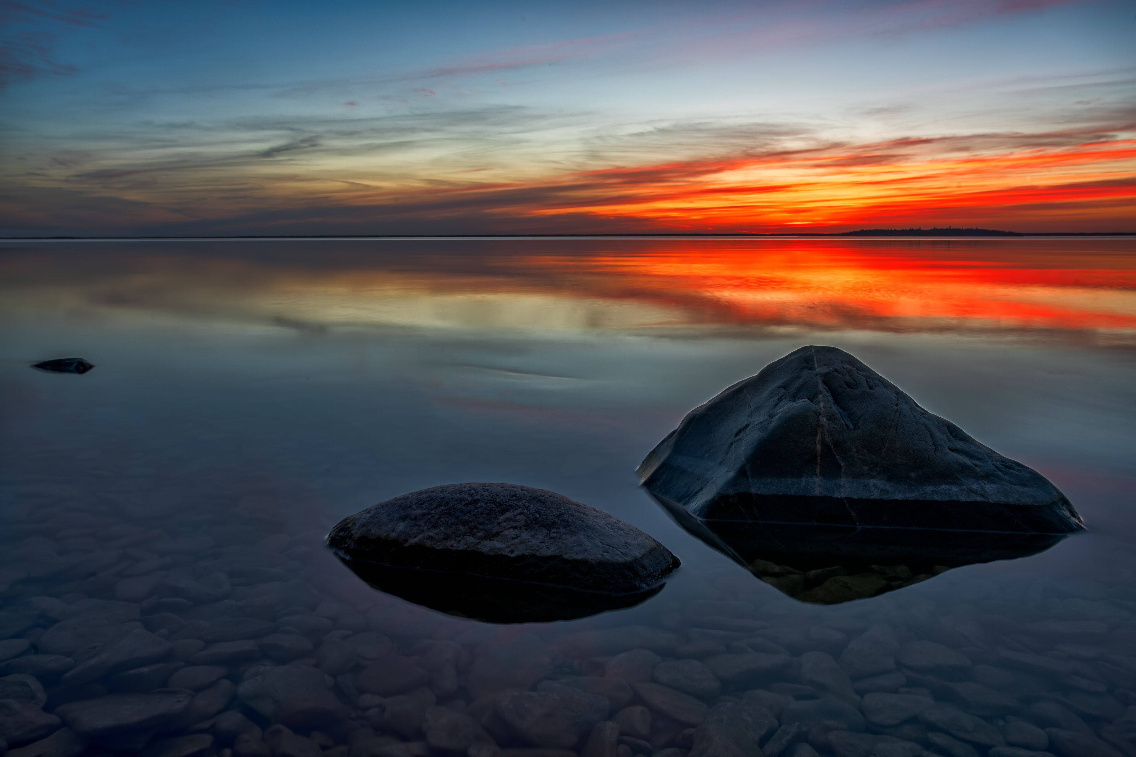 Um lago calmo com um pôr do sol, duas pedras grandes e um reflexo nítido do céu.