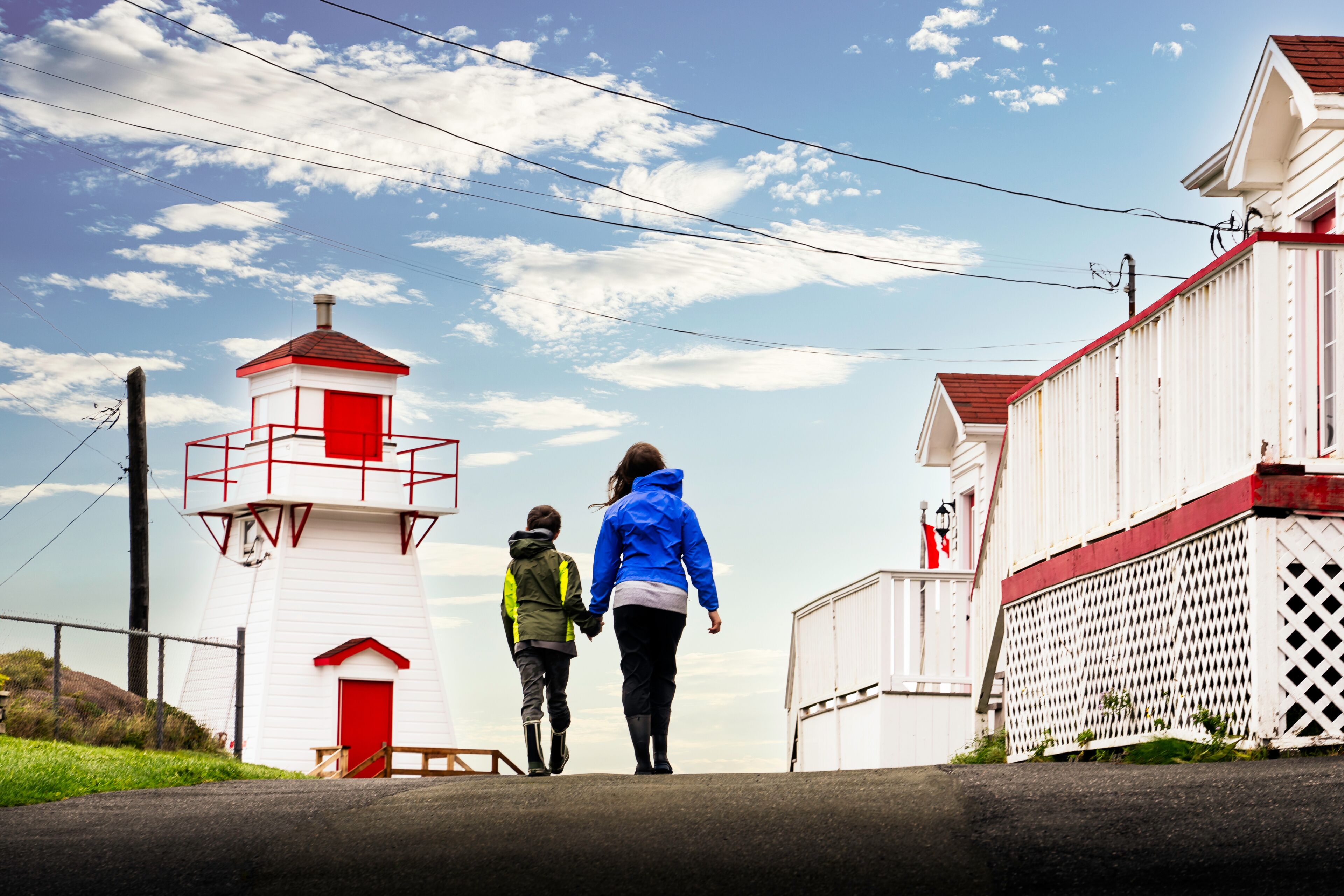 Um farol, uma pessoa com casaco azul e uma pessoa com casaco verde caminhando em direção ao farol.