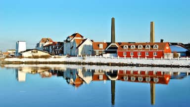 Uma vila à beira-mar com edifícios refletidos na água, uma chaminé alta e o chão coberto de neve.