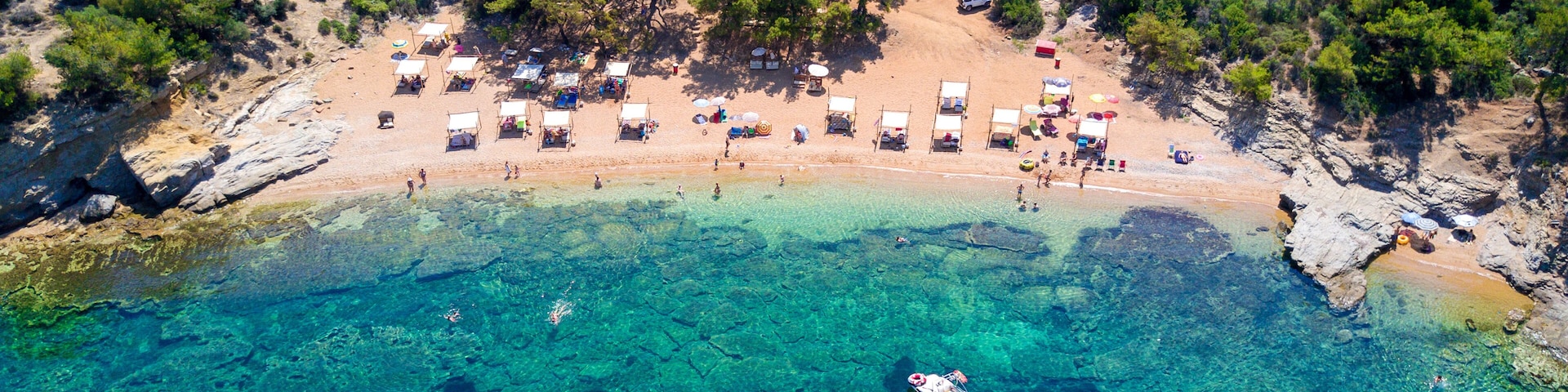Vista aérea de uma praia com águas turquesa cristalinas, um pequeno barco ancorado perto da costa e algumas cabanas e tendas de praia.