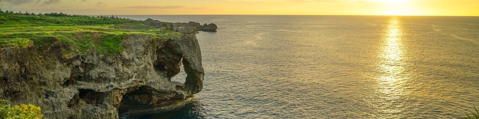 Paisagem costeira com uma formação rochosa imponente, apresentando um arco natural, vegetação exuberante e um pôr do sol sobre o oceano.