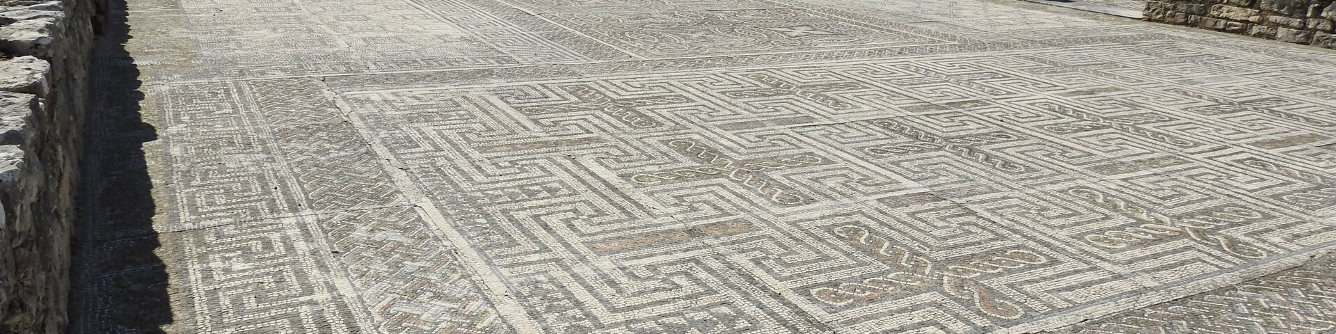 Um antigo chão de mosaico com padrões geométricos, cercado por paredes de pedra e ruínas.