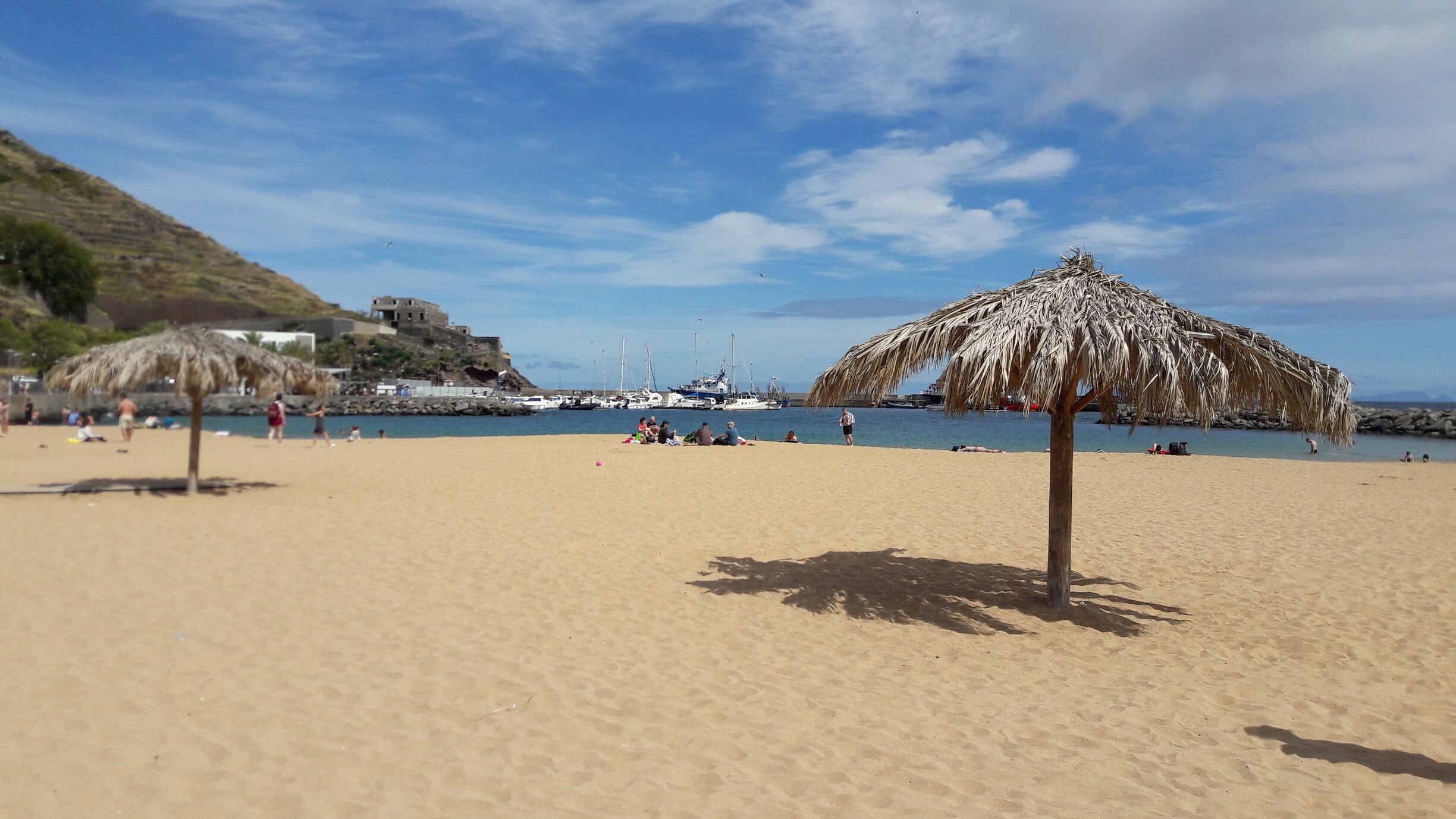 Uma praia com guarda-sóis de palha, pessoas relaxando e barcos na água.