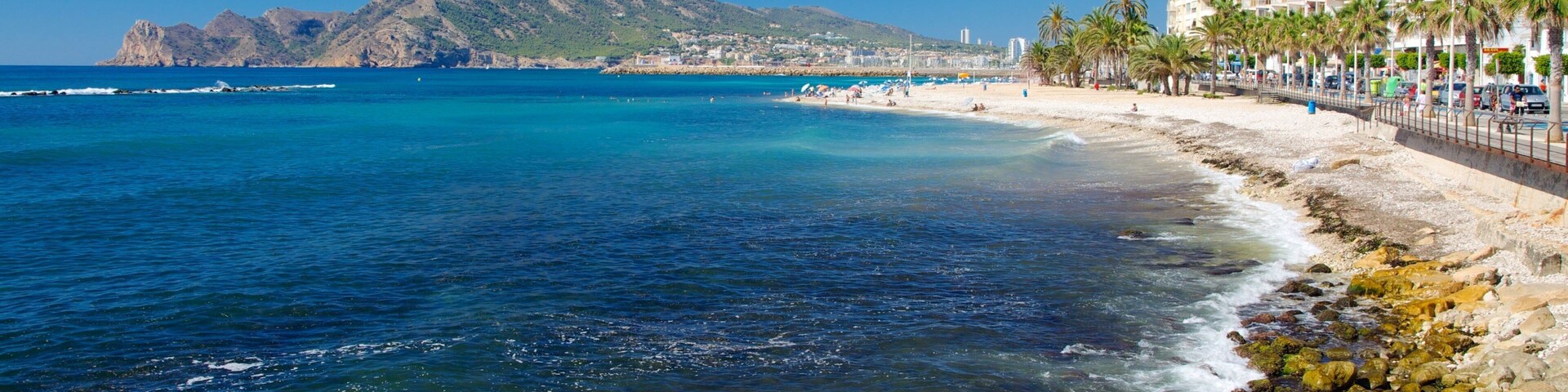 Uma praia costeira com águas azuis cristalinas, margem rochosa e uma montanha ao fundo.
