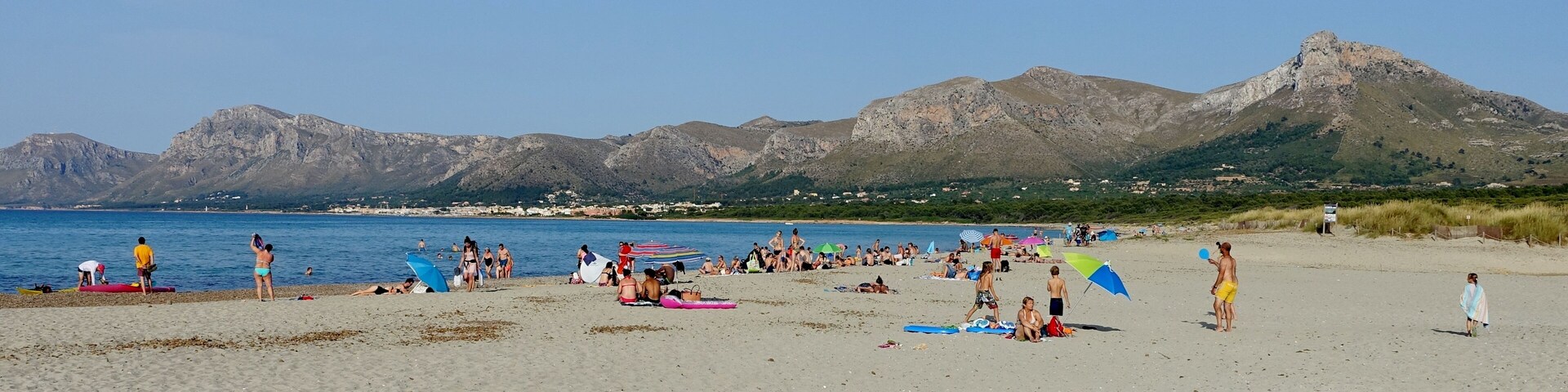 Uma praia com pessoas tomando sol e nadando, uma montanha ao fundo e um céu azul e límpido.