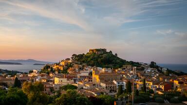 Uma vila costeira com um castelo no alto de uma colina, habitação densa e vista para o mar.