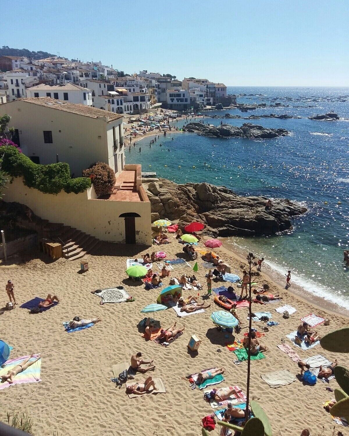Uma praia movimentada com pessoas se banhando ao sol e nadando, cercada por falésias rochosas e edifícios brancos.