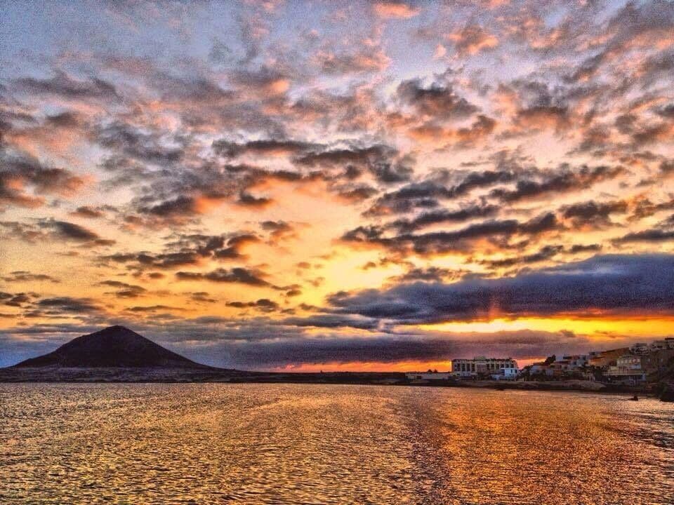 Pôr do sol sobre a água, com uma montanha ao fundo e edifícios na orla.